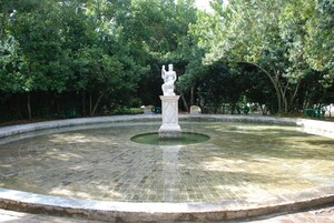 Uma fonte circular com uma estátua no centro, rodeada por um pavimento e árvores.