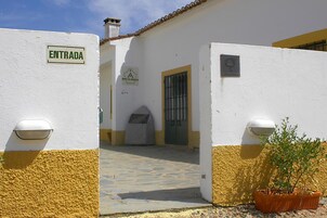 Um edifício branco com base amarela, uma placa de entrada, uma porta e uma pequena área de jardim com um vaso de planta.