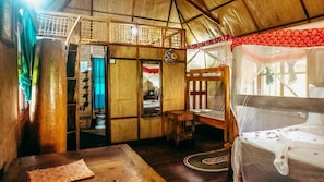 Quarto rústico com cama de madeira, uma pequena mesa e um banco. Há uma janela com cortina vermelha e mosquiteiro sobre a cama. Uma escada de madeira e uma prateleira pequena também são visíveis.