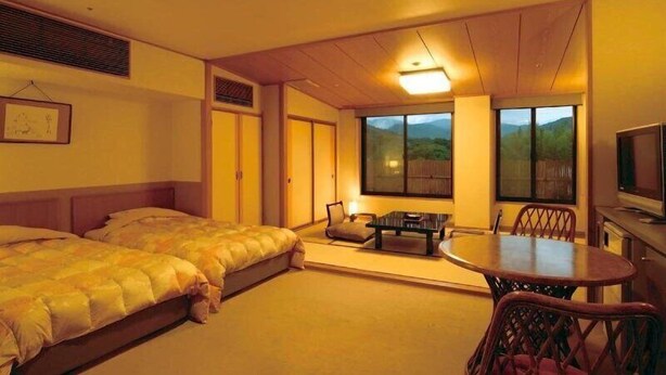Quarto de hotel com duas camas, uma mesa de jantar, cadeiras, uma TV e janelas amplas.