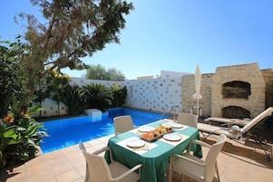 Uma área de refeições à beira da piscina, com uma mesa posta para dois, rodeada por cadeiras de lounge e uma parede de pedra com lareira.