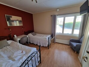 Quarto com duas camas, uma escrivaninha e uma cadeira. Há uma janela com cortinas, um radiador e um quadro na parede.
