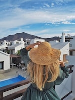Uma pessoa de cabelo loiro usando um chapéu de palha, em pé numa varanda com vista para uma piscina e edifícios brancos, com uma montanha ao fundo.