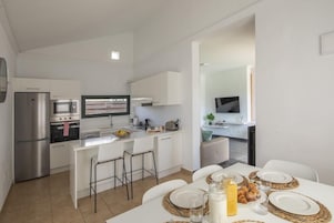 Cozinha moderna com armários brancos, geladeira de aço inoxidável, forno embutido e micro-ondas. Há uma área de jantar com uma mesa posta para dois, incluindo pratos, copos e um jarro de suco.