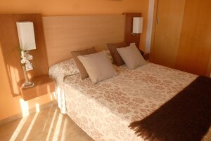 Cama bem arrumada com colcha estampada e vários travesseiros. Há uma mesa de cabeceira com luminária e um vaso com flores.