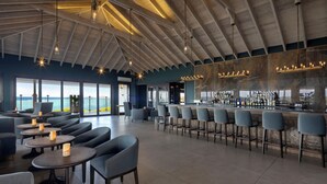 Uma área de bar ampla com diversas opções de assento, balcão repleto de garrafas e grandes janelas com vista para o mar.