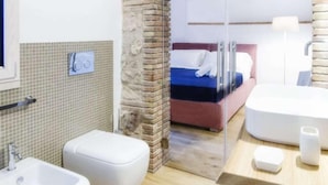 Casa de banho moderna com uma grande pia, sanita e uma cama visível através de um painel de vidro.