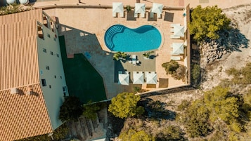 Vista aérea de um hotel com piscina, cercada por espreguiçadeiras e guarda-sóis, telhado de terracota e uma parede de pedra.