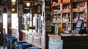 Um bar com banquetas de madeira, balcão de pedra e prateleiras repletas de garrafas diversas.
