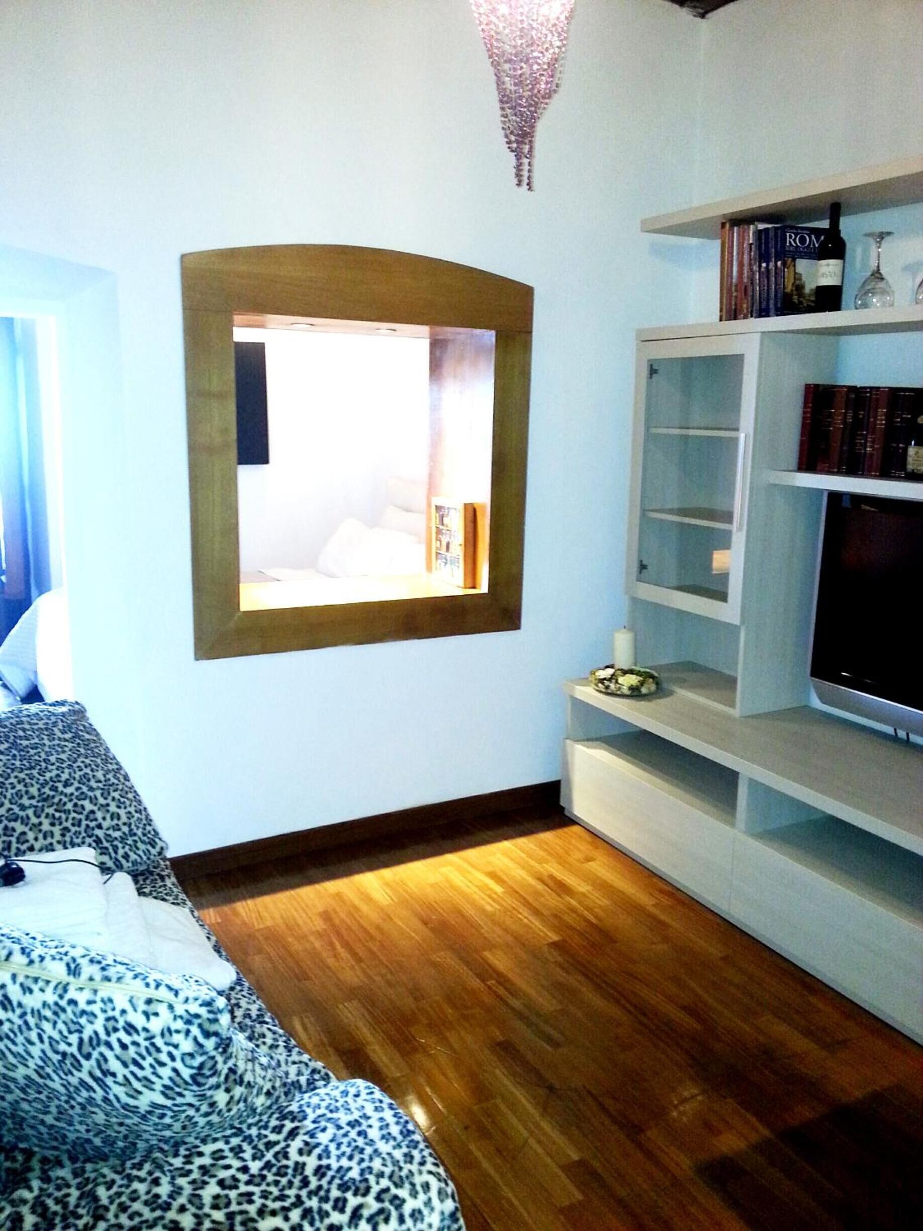 Uma sala de estar moderna com uma janela emoldurada em madeira, um rack branco com livros e uma TV, e um sofá com estampa de leopardo.