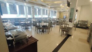 Uma sala de jantar ampla com várias mesas redondas e cadeiras, um buffet com estações de comida cobertas, e janelas grandes que permitem entrada de luz natural.