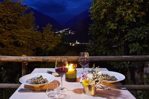 Mesa posta ao ar livre com pratos de comida, copos de vinho e uma vela, com vista para uma paisagem montanhosa.