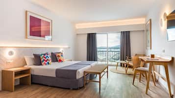 Quarto de hotel com cama, mesa de cabeceira, escrivaninha e cadeiras, janela grande com vista e quadro na parede.