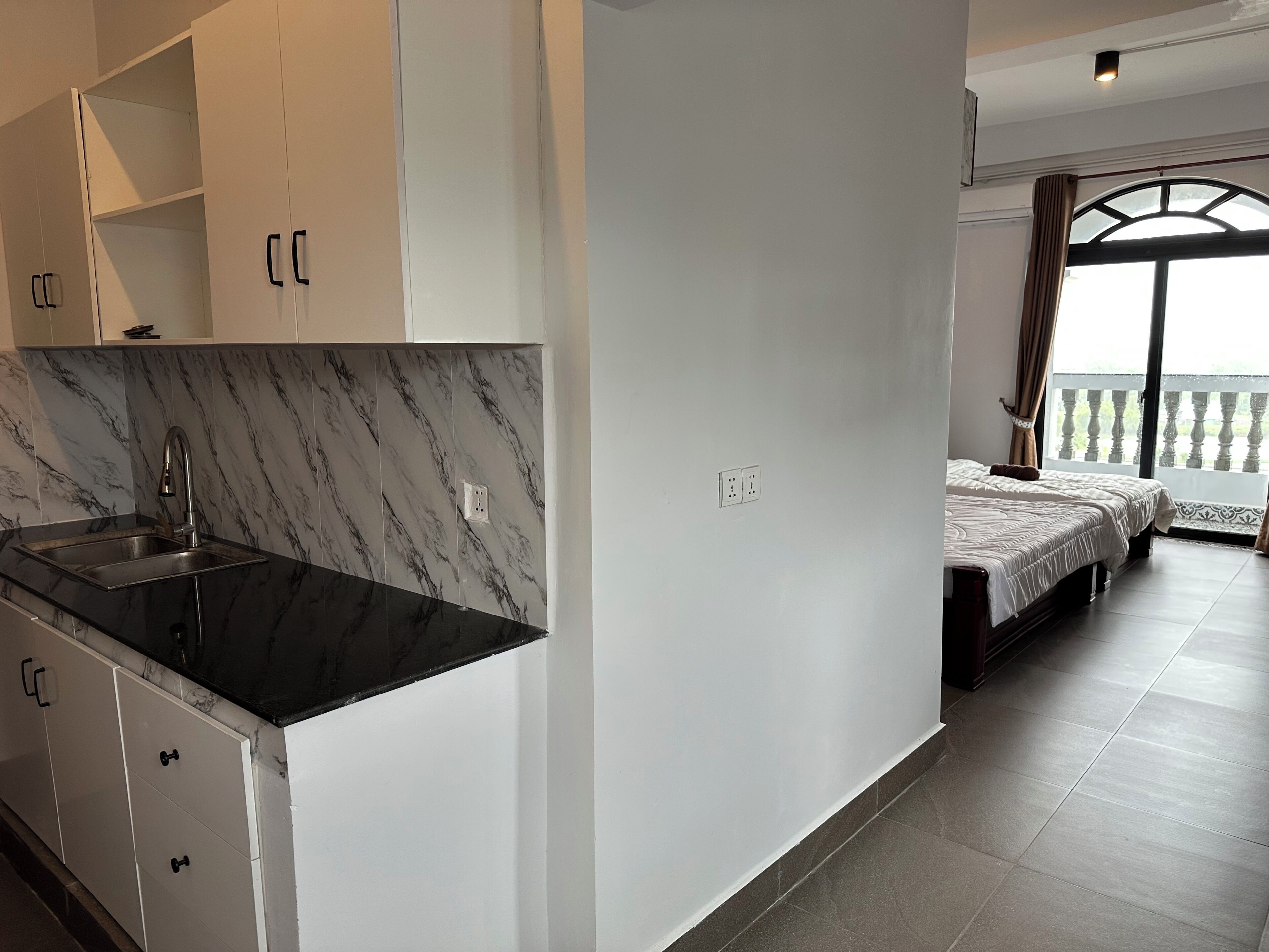 Cozinha moderna com bancadas de mármore e armários brancos, um quarto com vista para varanda e uma grande janela com cortinas.