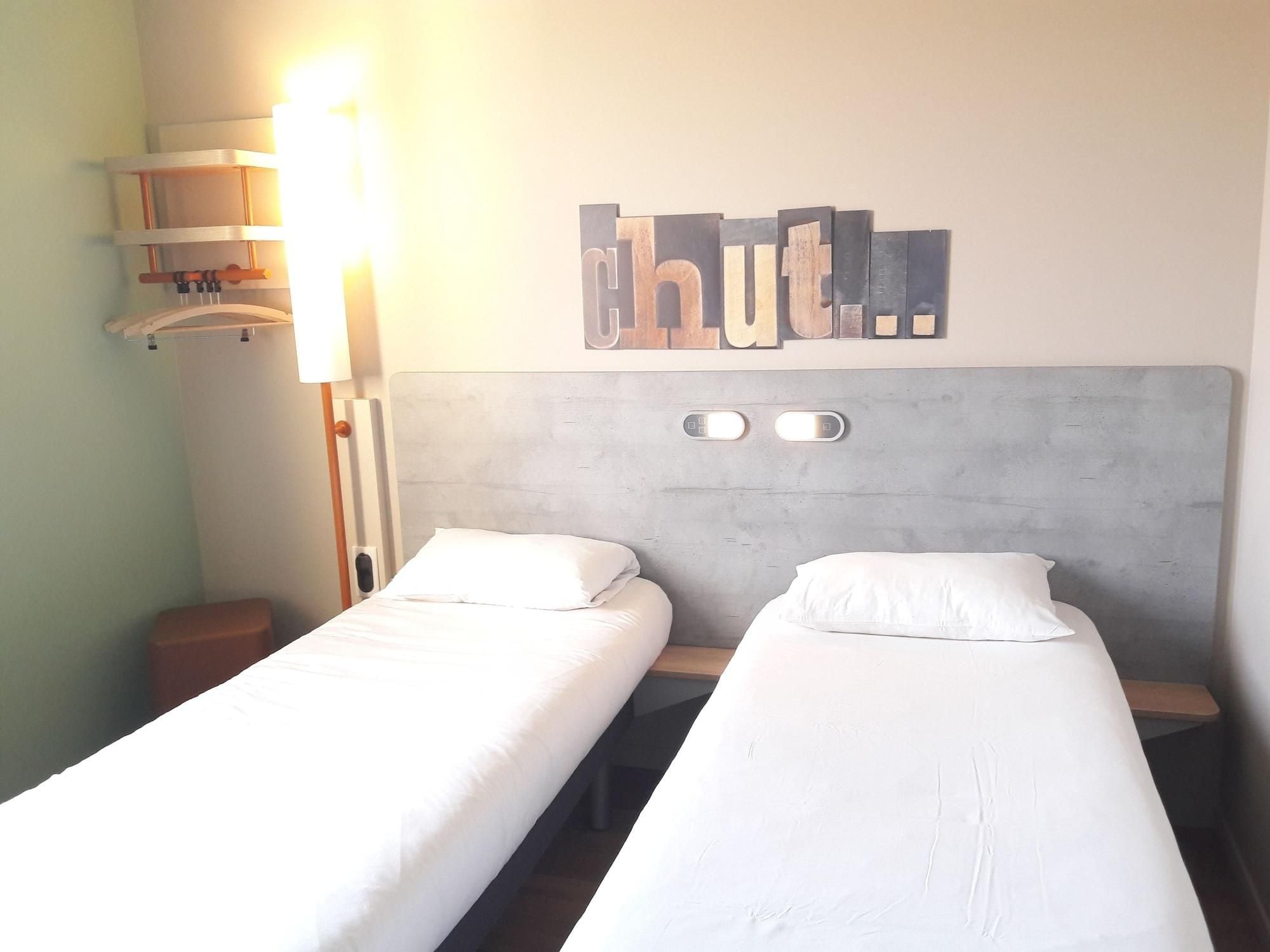 Quarto com duas camas, cabeceira, prateleira na parede e luminária de parede.