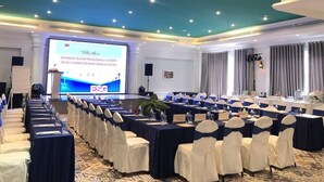 Uma sala de conferência com fileiras de mesas e cadeiras, uma tela grande exibindo uma apresentação e um palco com um púlpito.