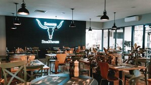 Interior de restaurante com um letreiro de neon que diz 'Seasons Steakhouse', mesas, cadeiras e luminárias pendentes.
