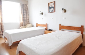 Quarto de hotel com duas camas de solteiro, uma mesa de cabeceira com abajur, um quadro na parede e uma janela com cortinas.