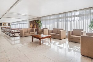 Um hall amplo com uma mesa de centro, vários sofás, uma escadaria e janelas grandes com cortinas leves.