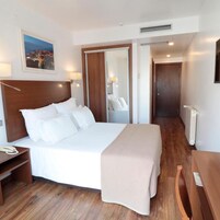 Quarto de hotel com uma cama grande, luminárias de cabeceira, um painel de madeira na cabeceira, telefone ao lado da cama, um banco de madeira, um radiador e uma porta que dá para outro quarto.