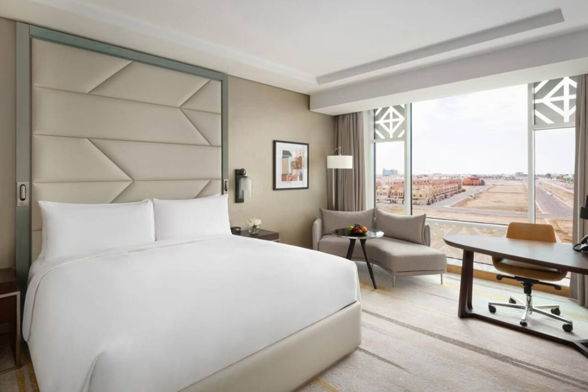Quarto de hotel com uma cama grande, uma escrivaninha, uma cadeira e vista para um canteiro de obras.