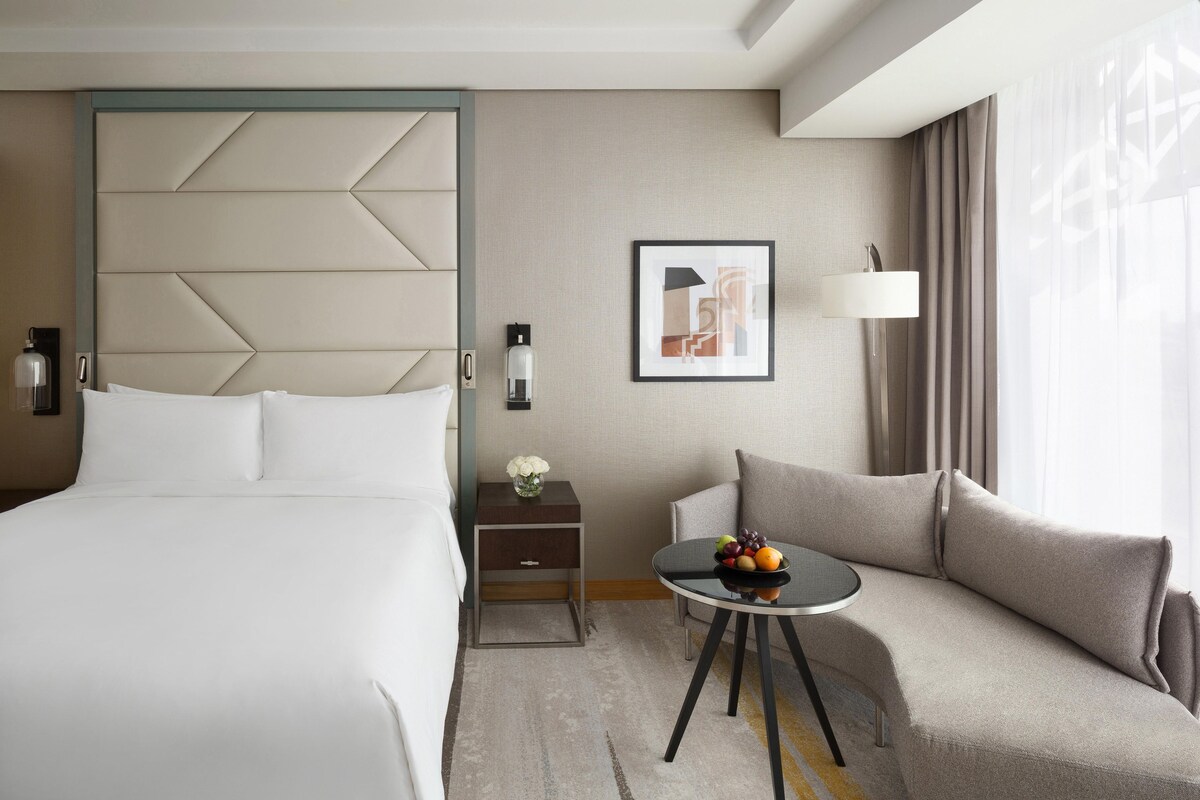 Quarto de hotel com cama, sofá, uma pequena mesa com frutas, mesa de cabeceira, abajur e um quadro na parede.