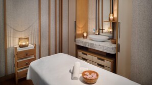 Quarto de hotel com cama, mesa de cabeceira de madeira, espelho, pia de mármore e uma tigela de madeira com flores.