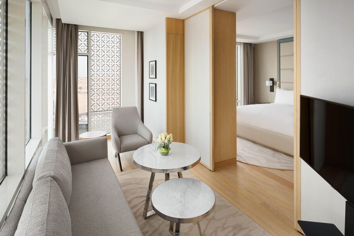 Quarto de hotel moderno com sofá, uma mesinha redonda com um vaso, cama e televisão.