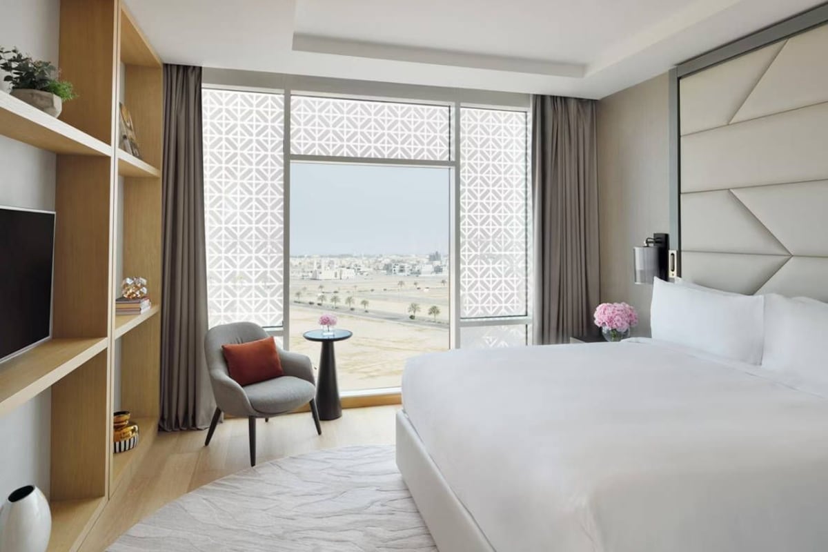Quarto de hotel com uma cama grande, televisão, cadeira, mesa pequena e vista para a cidade através de cortinas estampadas.