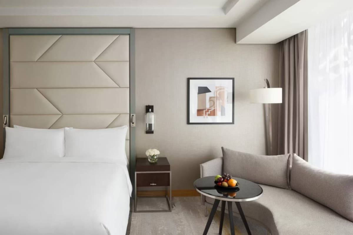 Quarto de hotel com cama, mesa de cabeceira, mesa com fruta, sofá, abajur e um quadro na parede.