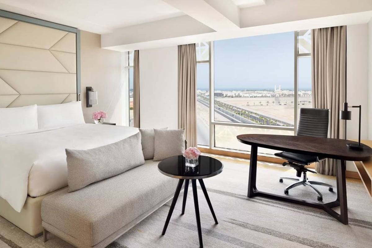 Quarto de hotel com cama, sofá, escrivaninha, cadeira e janelas amplas com vista para a cidade.