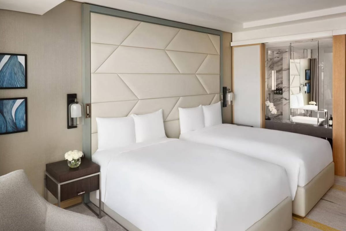 Quarto de hotel com duas camas, uma mesa de cabeceira, uma cadeira e um banheiro visível por meio de um separador de vidro.