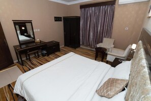 Quarto de hotel com uma cama grande, uma escrivaninha com espelho, uma cadeira, um guarda-roupa e uma cortina.