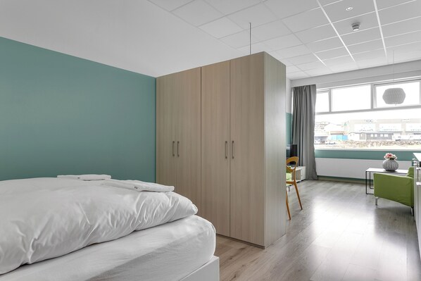Quarto moderno com uma cama grande, um guarda-roupa de madeira, uma pequena mesa com um vaso e vista para a cidade através de grandes janelas.