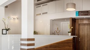 Uma área de recepção com balcão branco, parede de destaque em madeira e sinalização indicando comodidades como apartamentos, restaurante e praia.