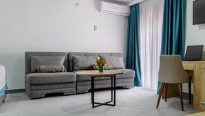 Quarto de hotel moderno com um sofá cinza, uma pequena mesa redonda com um vaso de flores, uma escrivaninha de madeira com um laptop e cortinas verde-água.