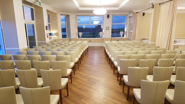 Uma sala de conferências com fileiras de cadeiras brancas, piso de madeira e janelas amplas com vista para o exterior.