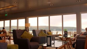 Um elegante hall de hotel com grandes janelas que oferecem vistas panorâmicas, assentos confortáveis e uma mesa de vidro.