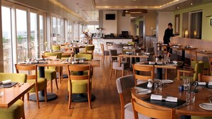 Um restaurante moderno com mesas e cadeiras de madeira, janelas grandes e um espaço de bar.