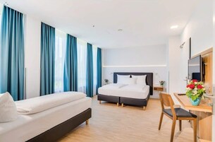 Quarto de hotel moderno com uma cama de solteiro, uma escrivaninha com cadeira e uma janela grande com cortinas.