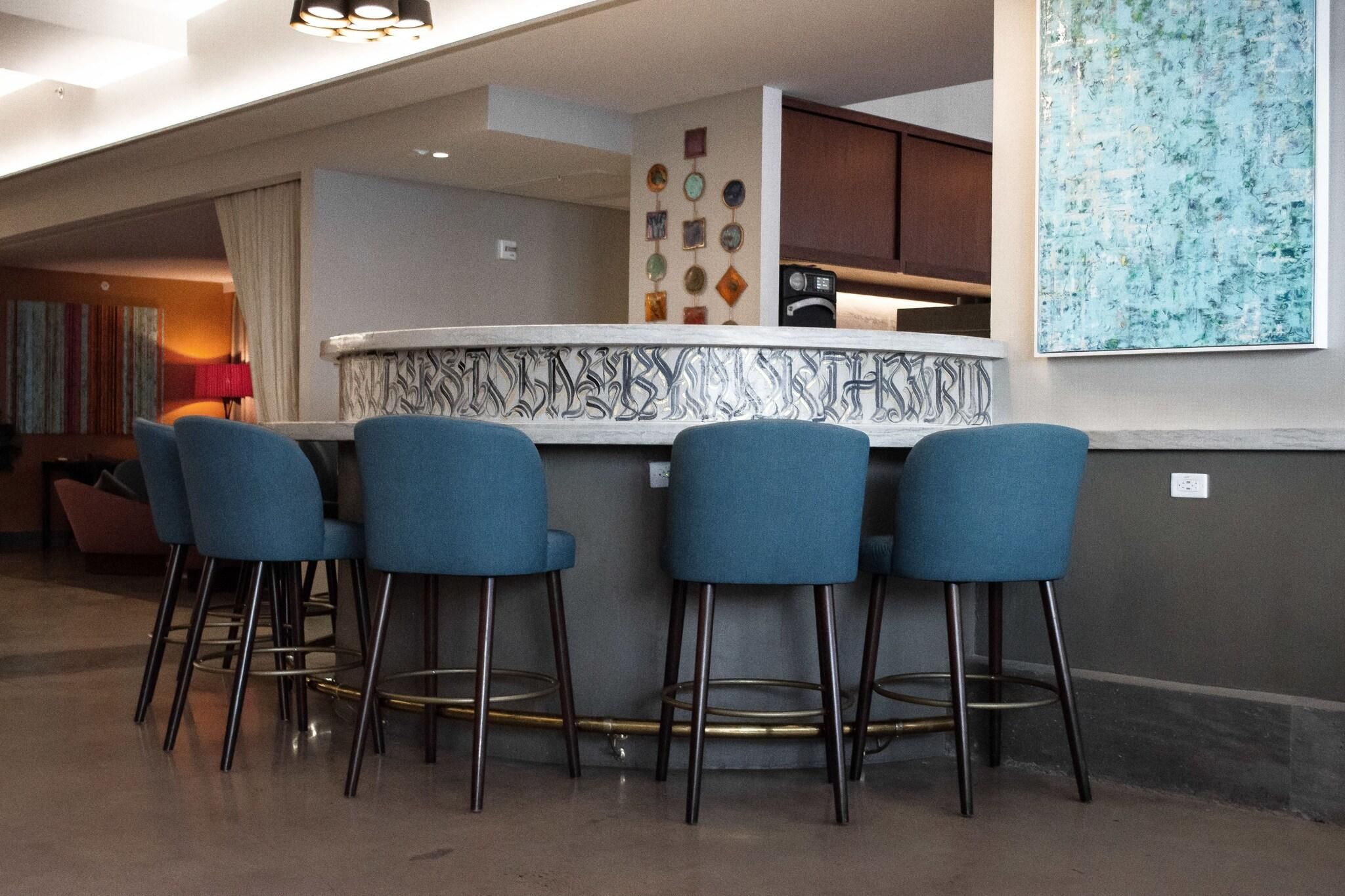 Uma área de bar com quatro banquetas azuis, um balcão de bar estampado e uma cozinha com armários e micro-ondas.