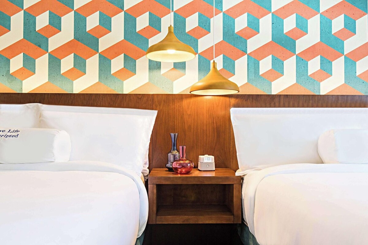 Quarto de hotel com duas camas, uma cabeceira de madeira, uma mesa de cabeceira com abajur e uma parede com padrão geométrico.