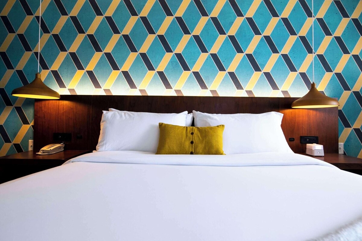 Quarto de hotel com parede estampada, cama com roupa de cama branca e amarela, e duas luminárias de cabeceira.