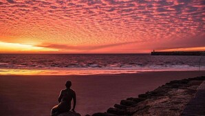 Um pôr do sol sobre uma praia, com a silhueta de uma pessoa sentada em uma rocha.