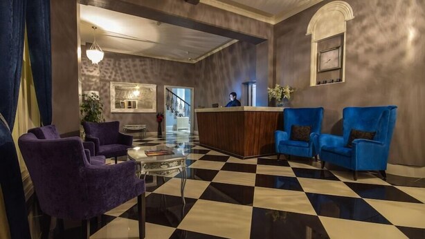 Um hall com piso xadrez, poltronas roxas e azuis, um balcão de recepção e uma escada ao fundo.