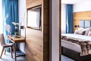 Quarto de hotel com escrivaninha de madeira, uma cadeira, cama com cabeceira cinza e uma janela grande com cortinas.