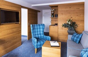 Um quarto com uma poltrona azul xadrez, um sofá cinza, uma mesa de centro de madeira e uma TV fixada em uma parede de madeira.