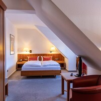 Quarto de hotel com teto inclinado, cama com lençóis brancos, duas luminárias de cabeceira, mesa de cabeceira de madeira, televisão e uma cadeira vermelha.