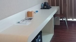 Quarto de hotel com uma escrivaninha branca, televisão fixada na parede, janela com cortinas e chão de madeira escura.