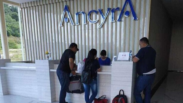 Um balcão de recepção com uma placa indicando "Ancyra" em um saguão moderno com piso de mármore e persianas verticais.